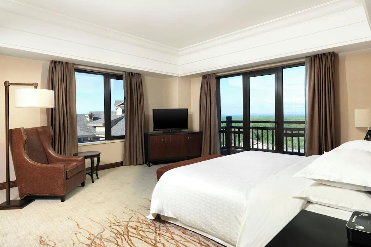 Quarto de hotel com uma cama grande, televisão, cadeira, mesinha e varanda com vista.