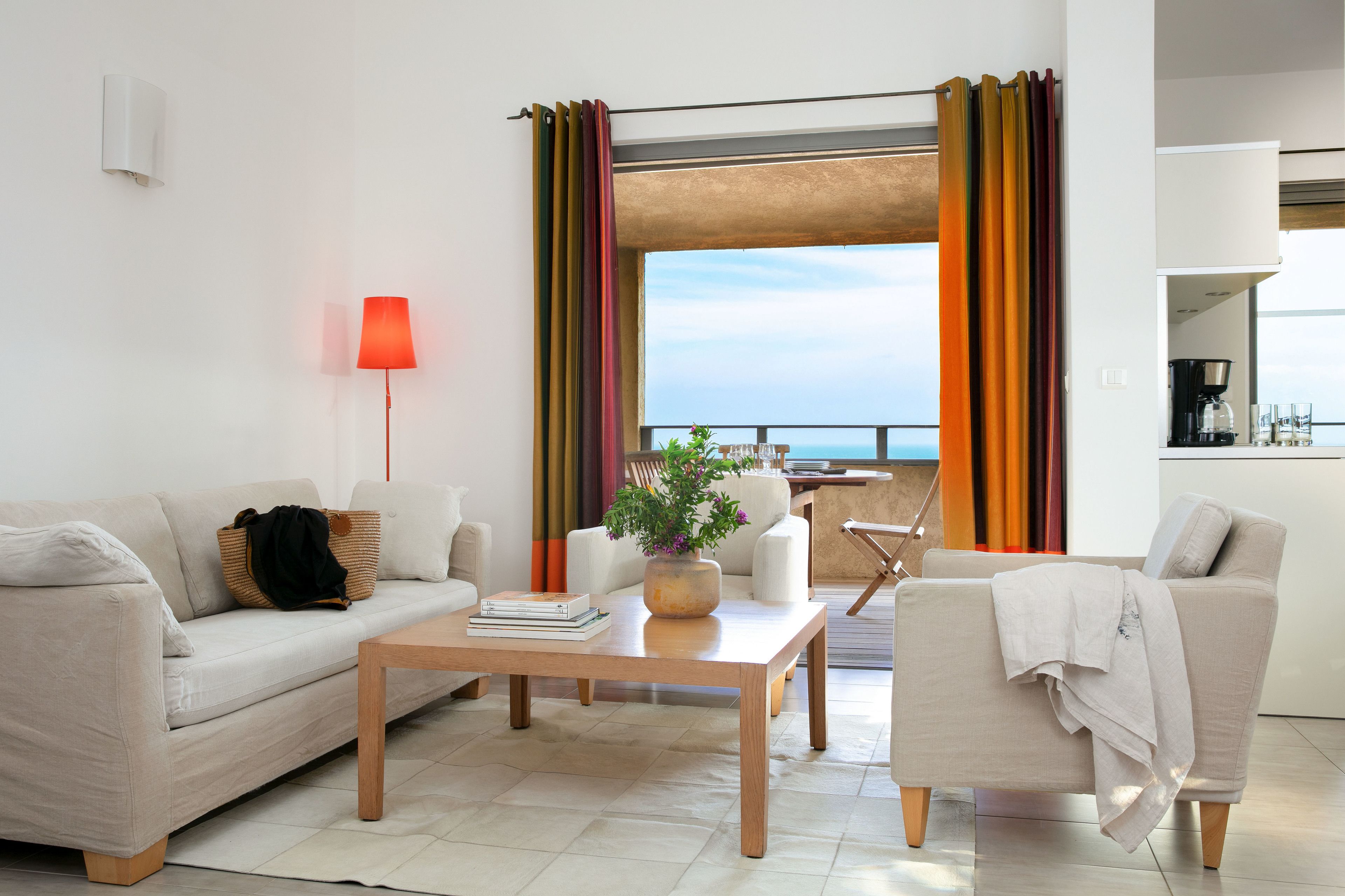 Uma sala de estar moderna com sofá, poltrona e mesa de centro, vista para o mar e área de cozinha.
