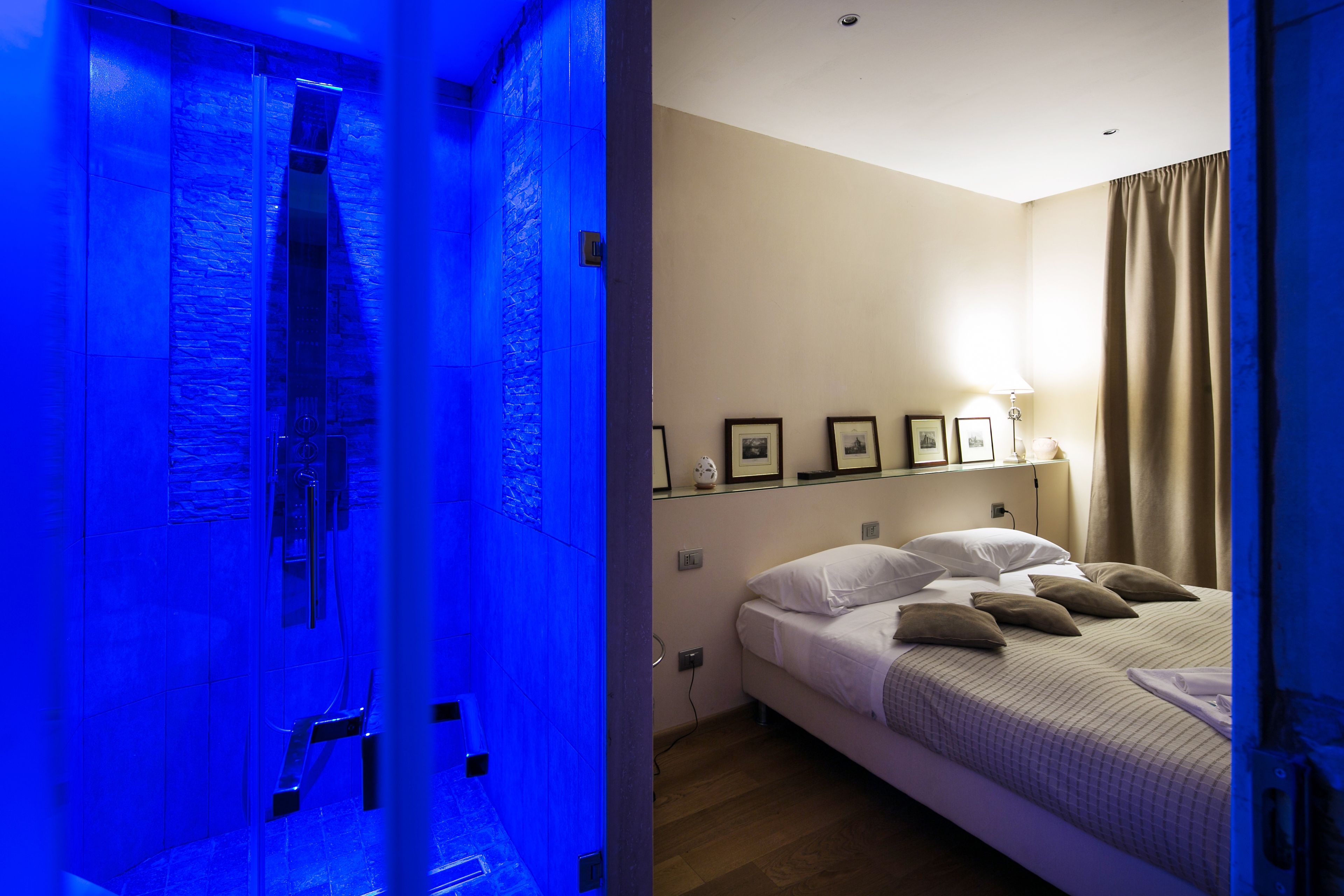 Quarto de hotel com uma cama, uma mesa de cabeceira com fotografias emolduradas e um banheiro com iluminação azul.