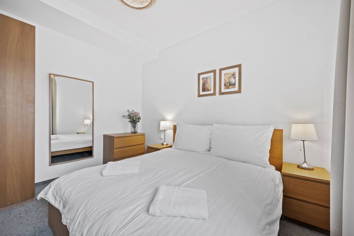 Quarto de hotel com uma cama grande, uma mesinha de cabeceira de madeira, uma mesa de apoio com abajur, um espelho e duas fotos emolduradas na parede.
