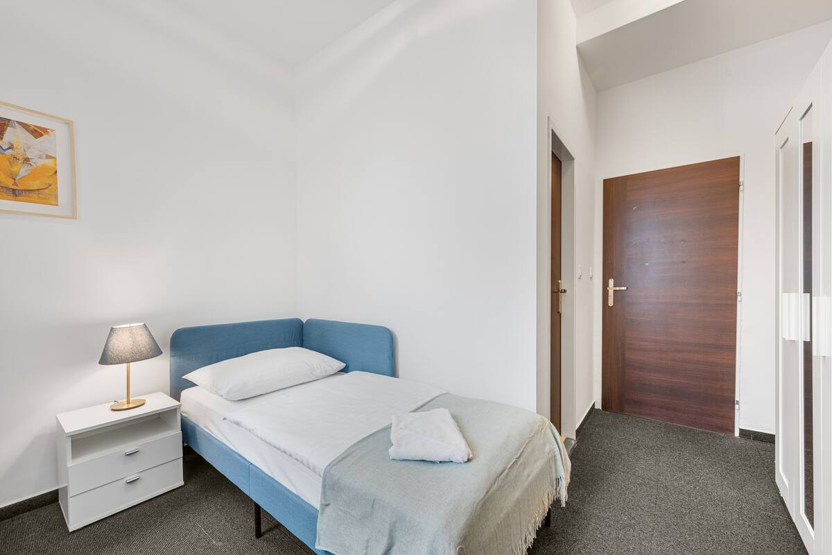 Um quarto com cama, mesa de cabeceira com luminária, porta e parede com quadro.