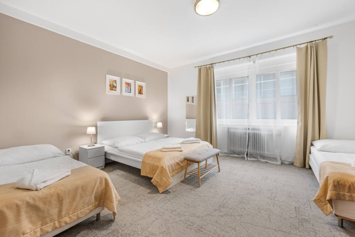 Quarto de hotel com duas camas, uma mesa de cabeceira com abajur, uma janela com cortinas e uma obra de arte na parede.