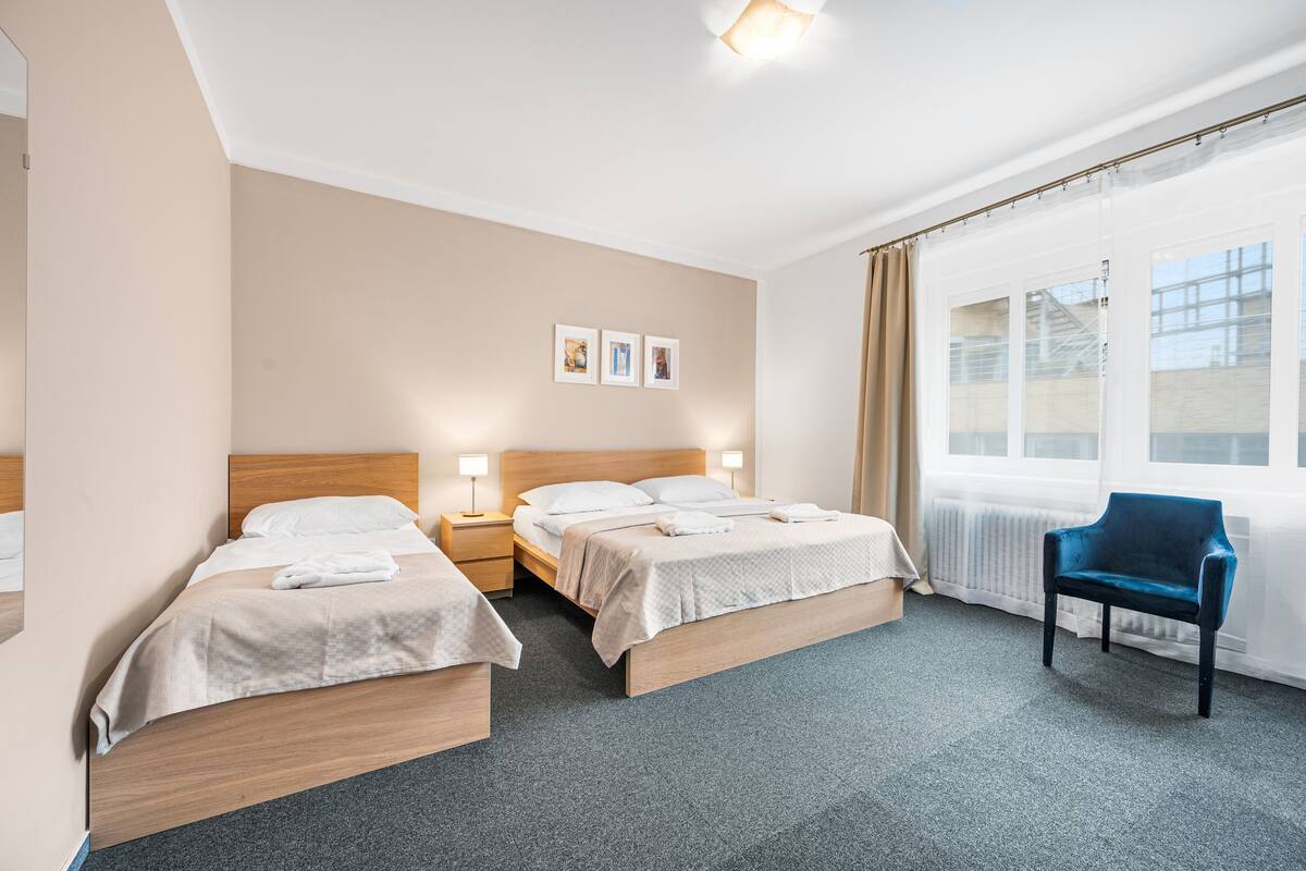 Quarto de hotel com duas camas, uma cadeira azul e uma janela grande com vista para um edifício.