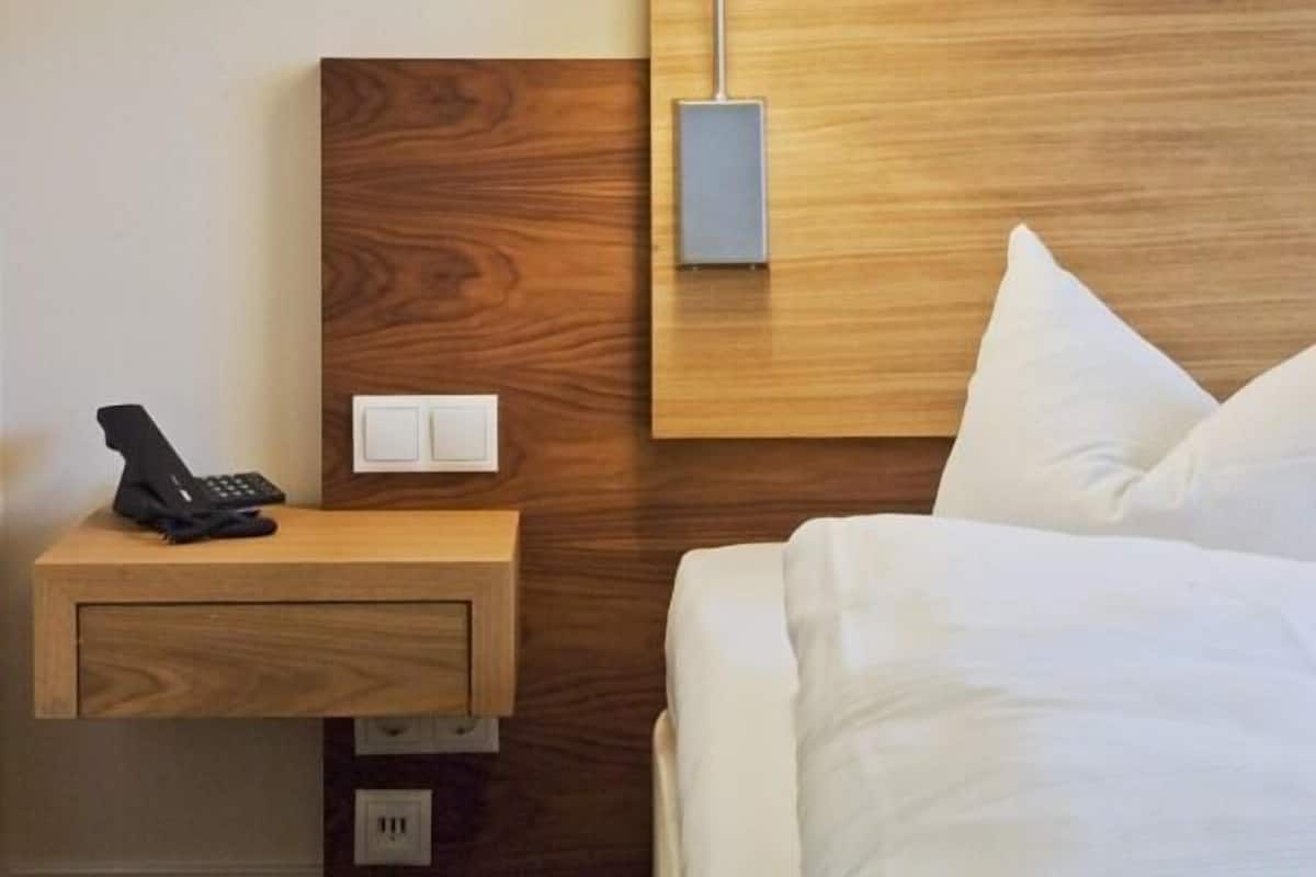 Quarto de hotel com cabeceira de madeira, mesa de cabeceira com abajur, telefone e tomada.