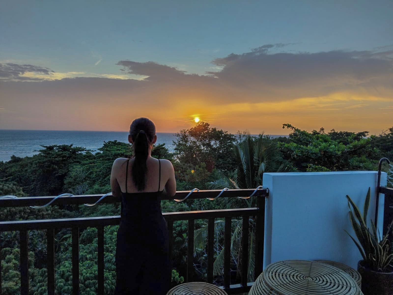 Uma pessoa está em uma varanda observando o pôr do sol, com vista para o oceano e vegetação densa.