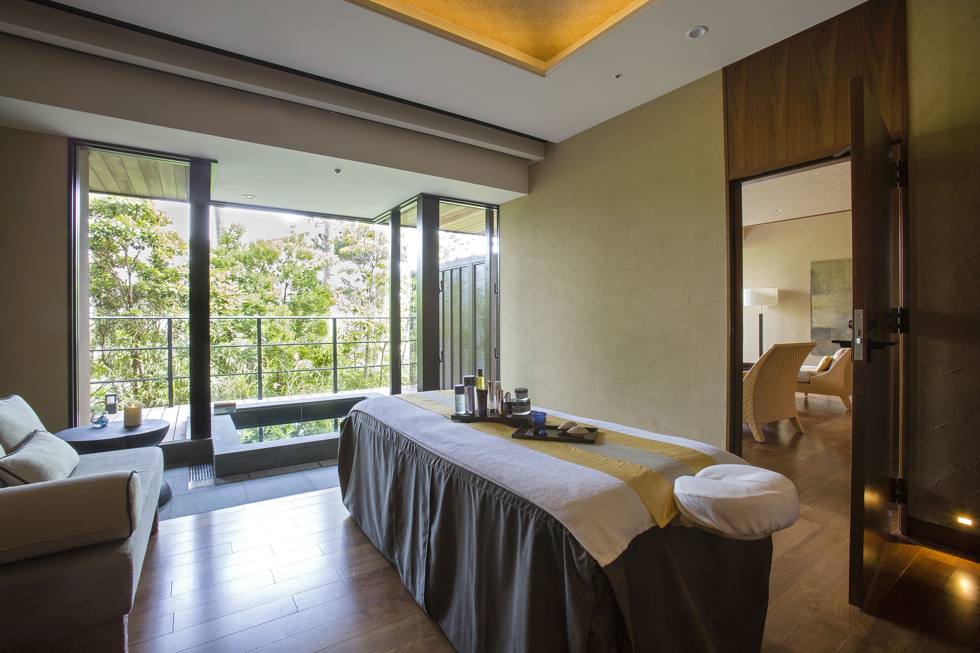 Quarto de hotel moderno com uma cama grande, um sofá, uma mesa de massagem e vista para o verde através de amplas janelas.