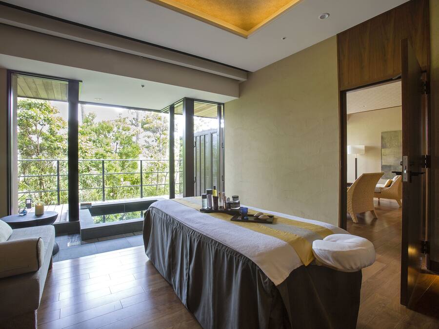 Quarto de hotel moderno com uma cama grande, um sofá, uma mesa de massagem e vista para o verde através de amplas janelas.