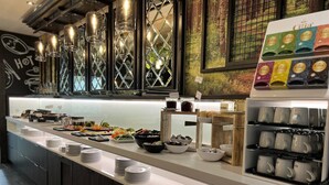 Uma mesa de buffet com diversos alimentos e acompanhamentos, um espaço para café e uma parede com um mural de floresta.