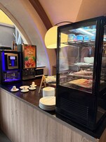 Uma estação de café com uma máquina de café KREa, uma vitrine de vidro com doces e uma placa com o texto 'Les Vergeres de Philomène'.