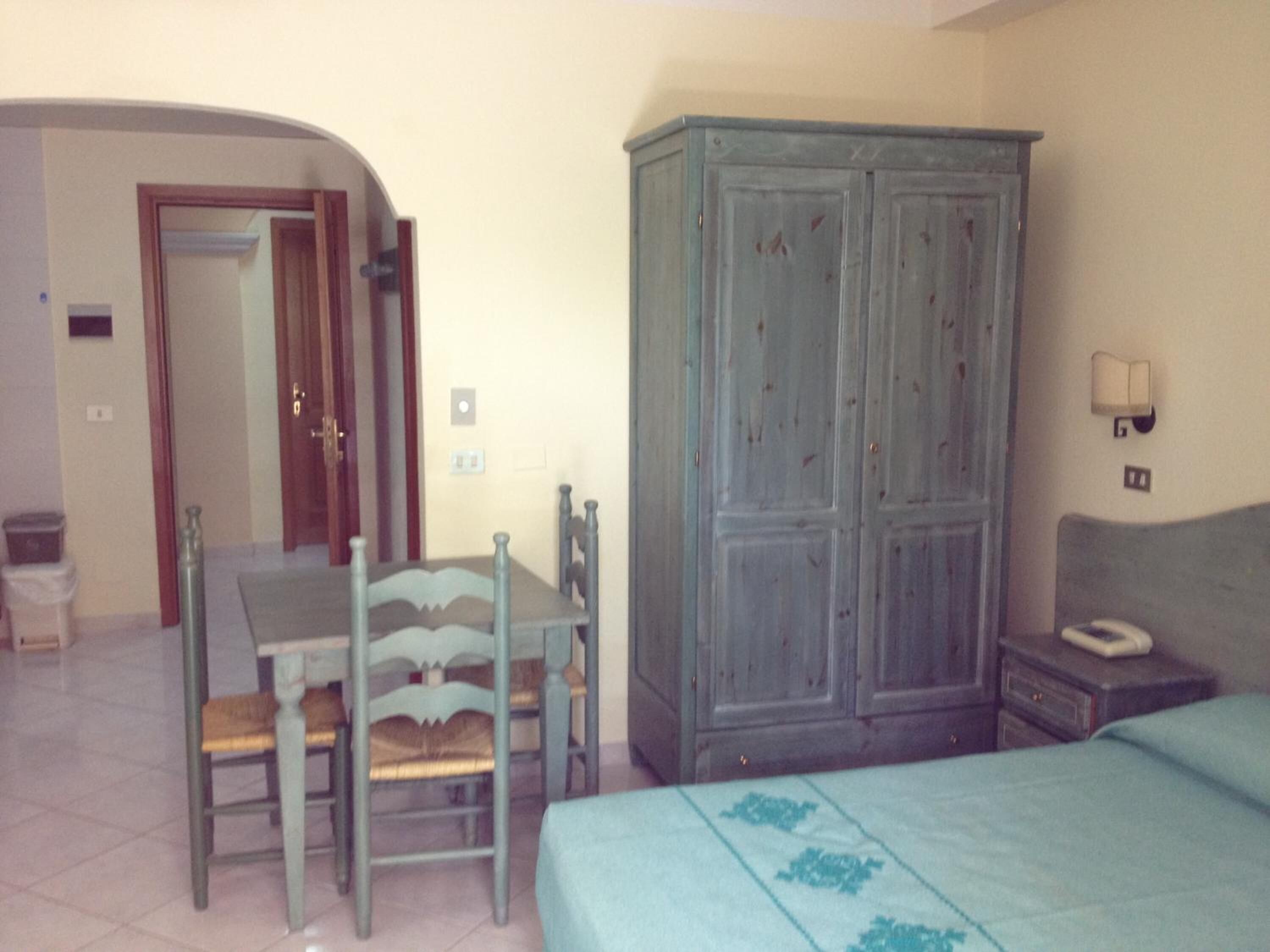Quarto com cama, mesa e cadeiras, guarda-roupa e porta que dá para outro cômodo.