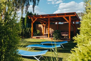 Uma pérgola de madeira com cobertura, um espaço de estar coberto e duas espreguiçadeiras azuis sobre um gramado.