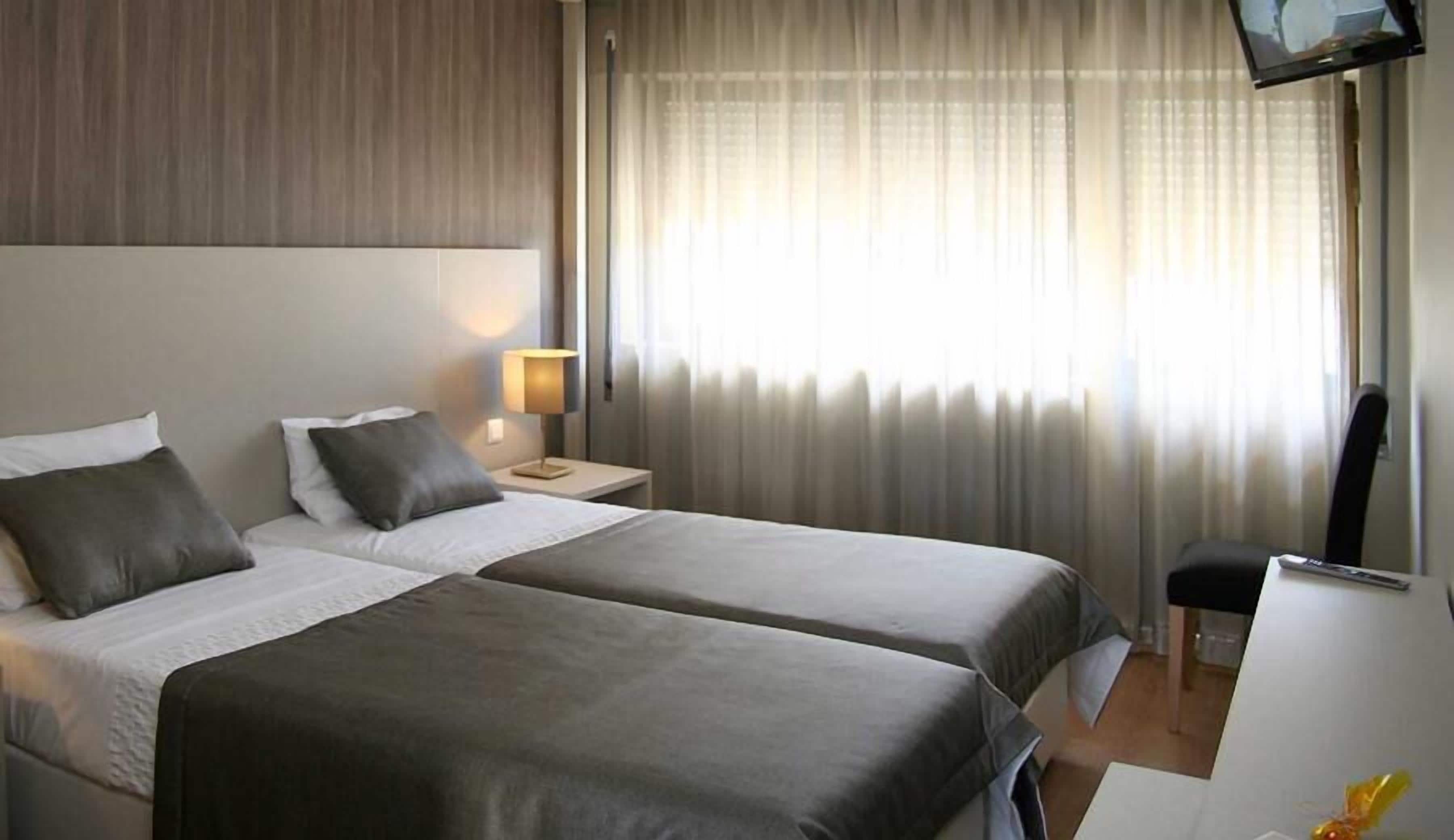 Quarto de hotel com duas camas, uma escrivaninha, uma cadeira, uma televisão e uma janela com cortinas.