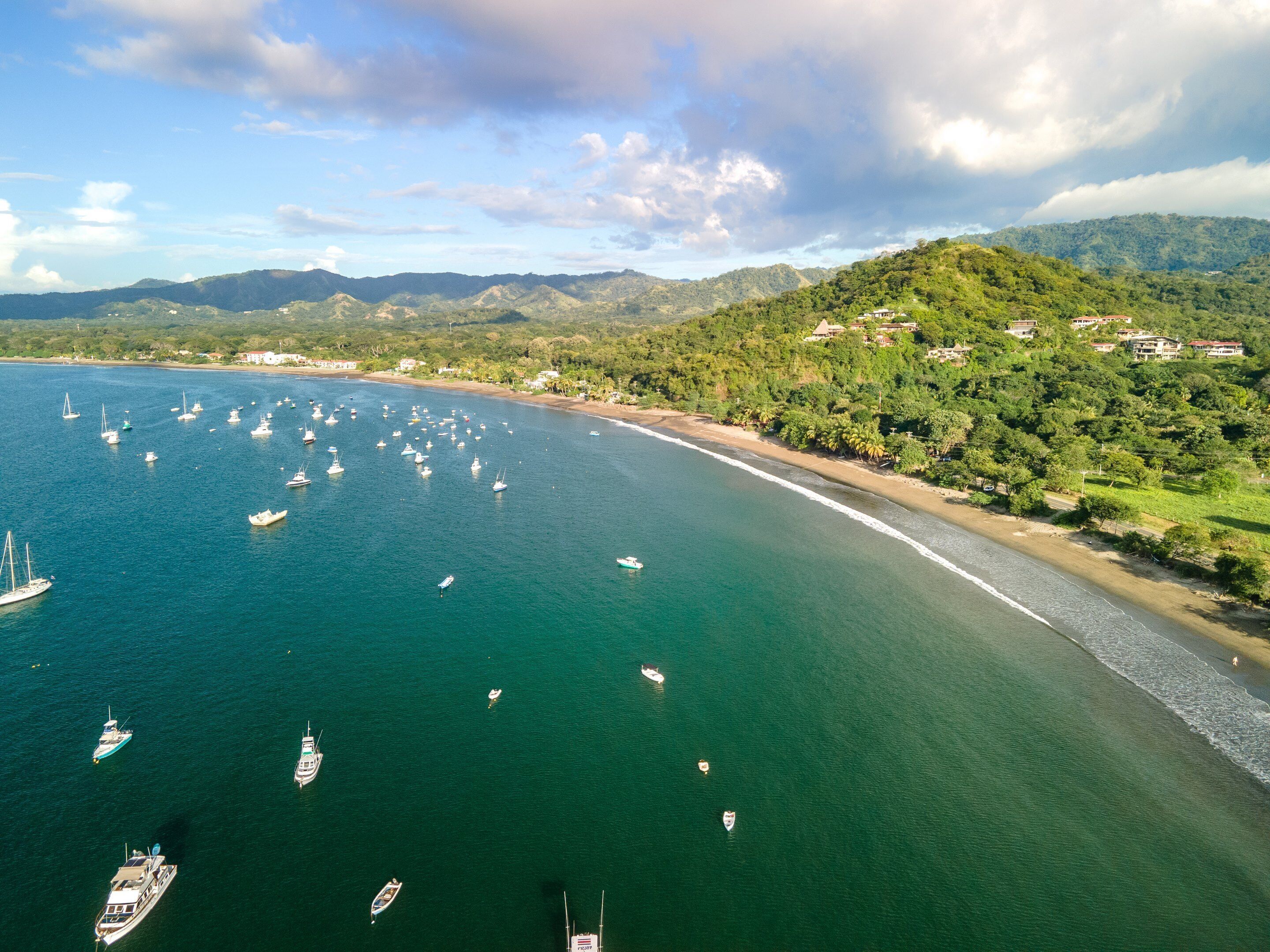 Vista aérea de uma área costeira com vários barcos na água, uma praia e colinas ao fundo.