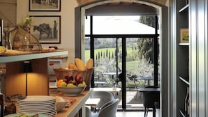 Uma cozinha com piso de ladrilhos em padrão herringbone, um balcão de café da manhã com frutas e pão, e vista para um jardim através de uma porta de vidro.