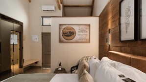 Quarto de hotel com uma cama, uma placa histórica emoldurada, uma pequena mesa com uma garrafa e um aparelho de ar condicionado fixado na parede.