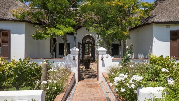 Uma trilha leva a um edifício tradicional com telhado de palha, cercado por vegetação exuberante e paredes brancas.