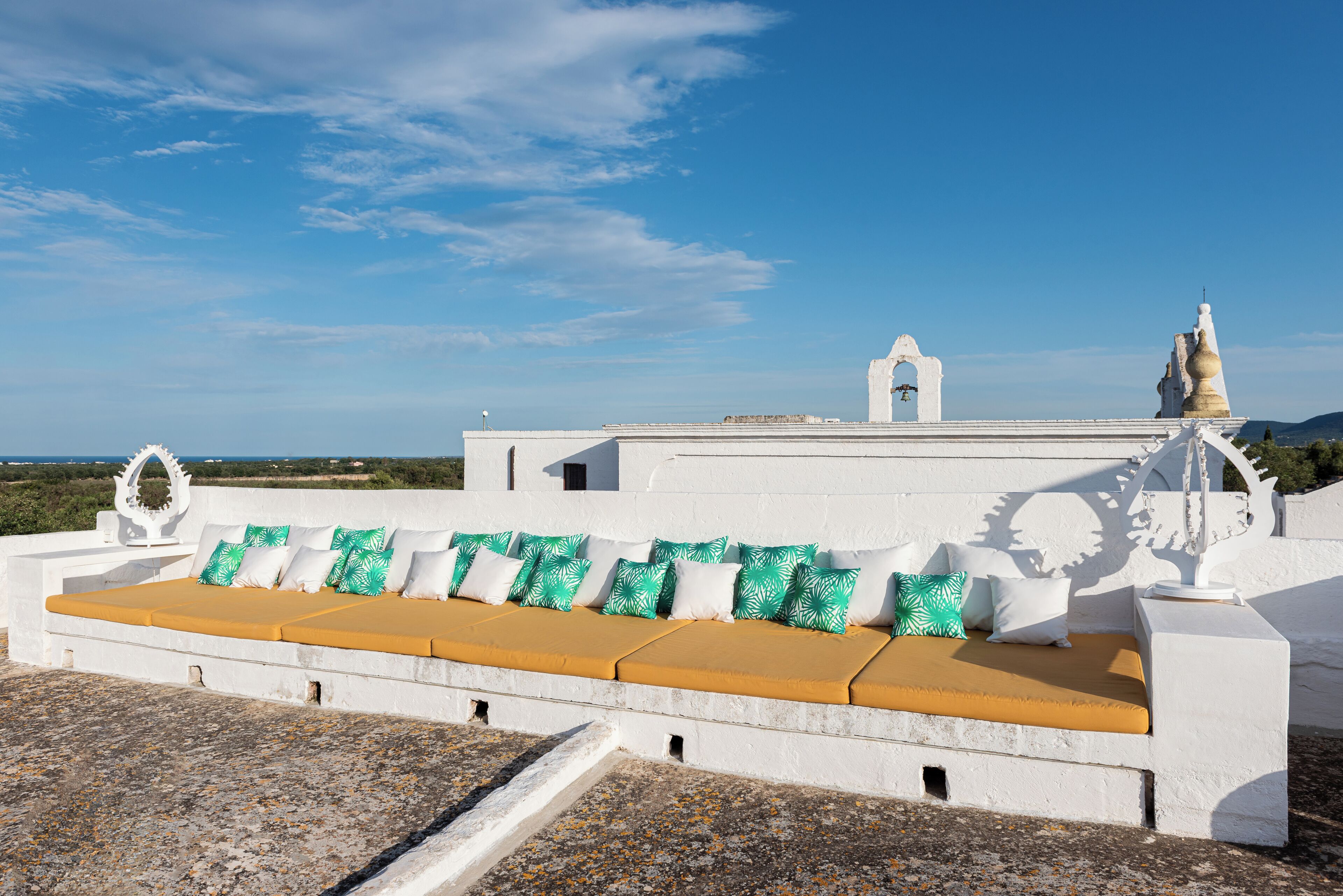 Terraço no telhado com uma parede branca, um banco acolchoado amarelo e uma escultura branca decorativa.