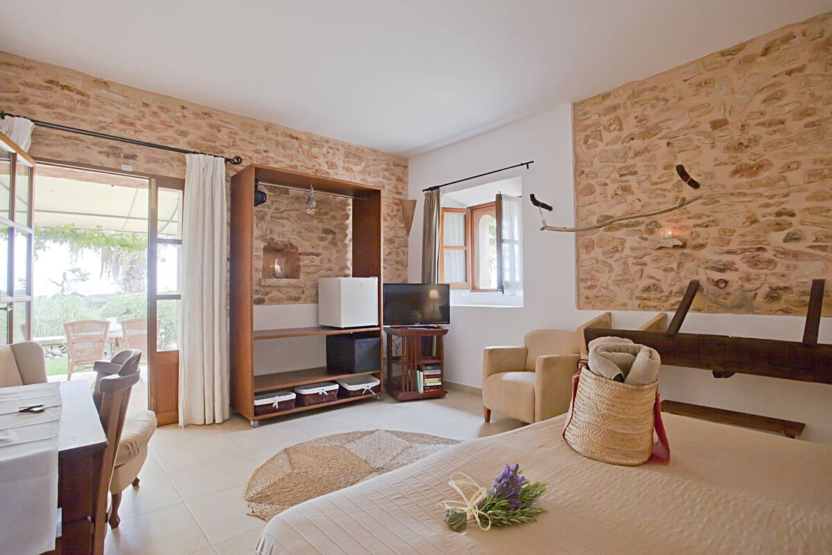 Quarto com parede de pedra, cama com um arranjo floral, TV, prateleira de madeira e área de jantar com cadeiras.