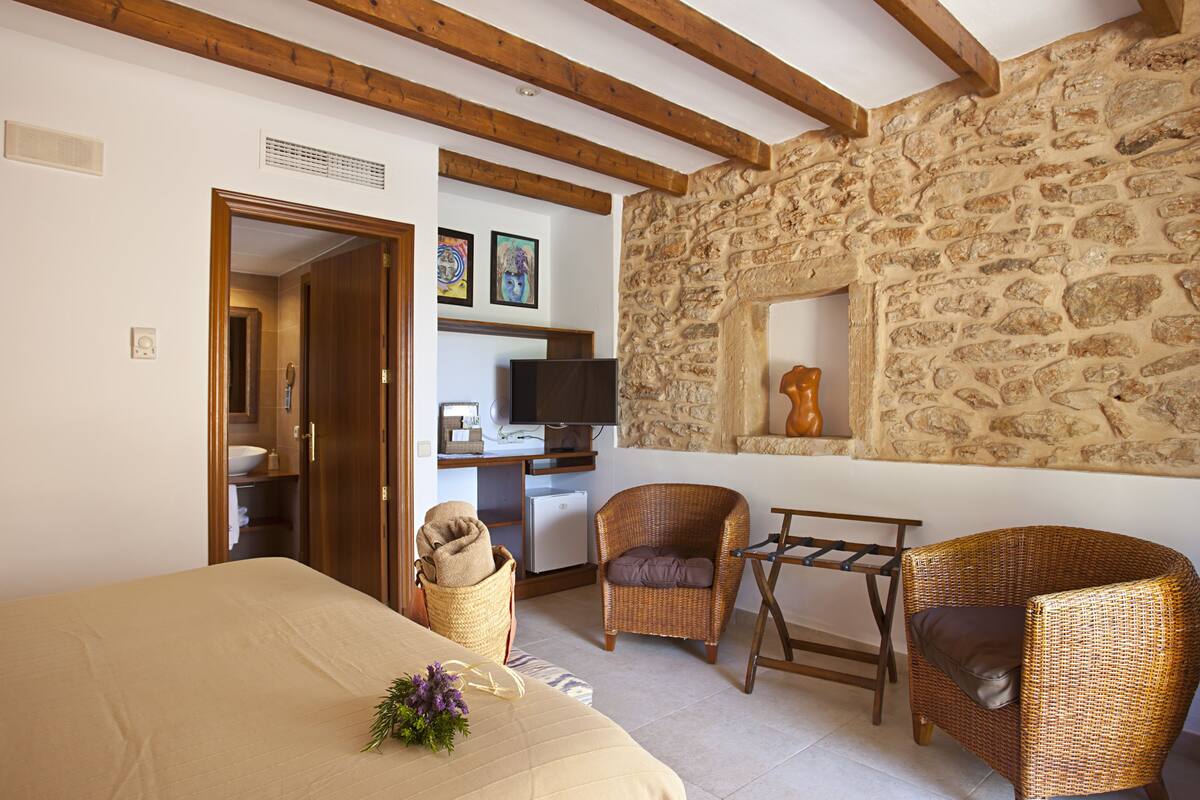 Quarto com cama, cadeiras de vime, uma mesa pequena, parede de pedra, televisão e banheiro visível por uma porta aberta.