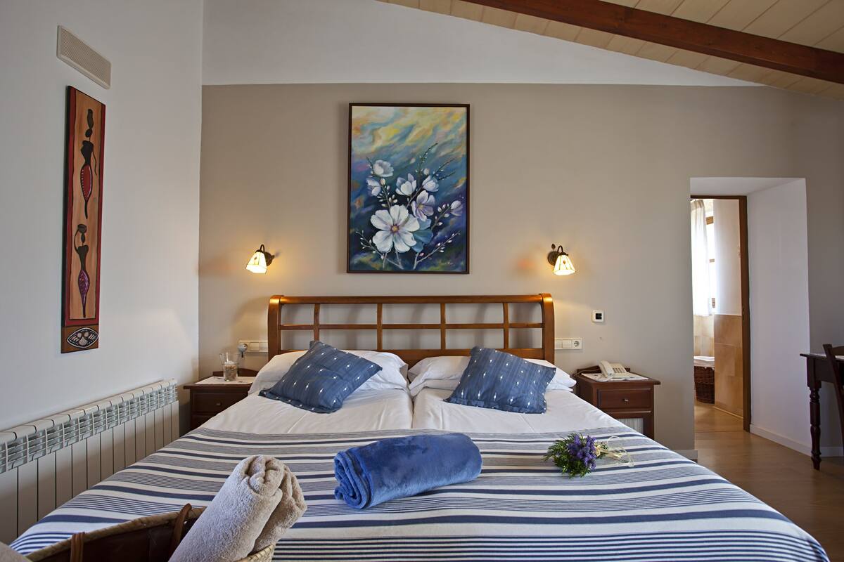 Quarto de hotel com uma cama grande, duas mesas de cabeceira, um quadro sobre a cama e vista para o corredor.