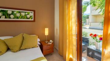 Quarto de hotel com cama, mesa de cabeceira, abajur e vista para uma varanda externa florida.