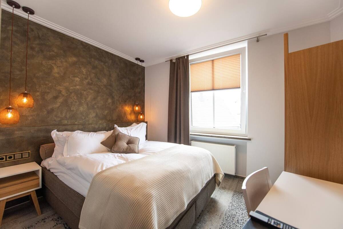 Quarto de hotel moderno com cama grande, mesas de cabeceira, escrivaninha e janela com persianas.