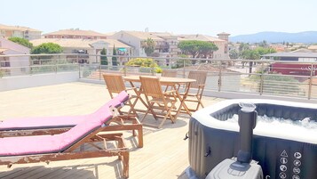 Terraço no telhado com cadeiras de lounge de madeira, hidromassagem e vista para edifícios residenciais e montanhas.