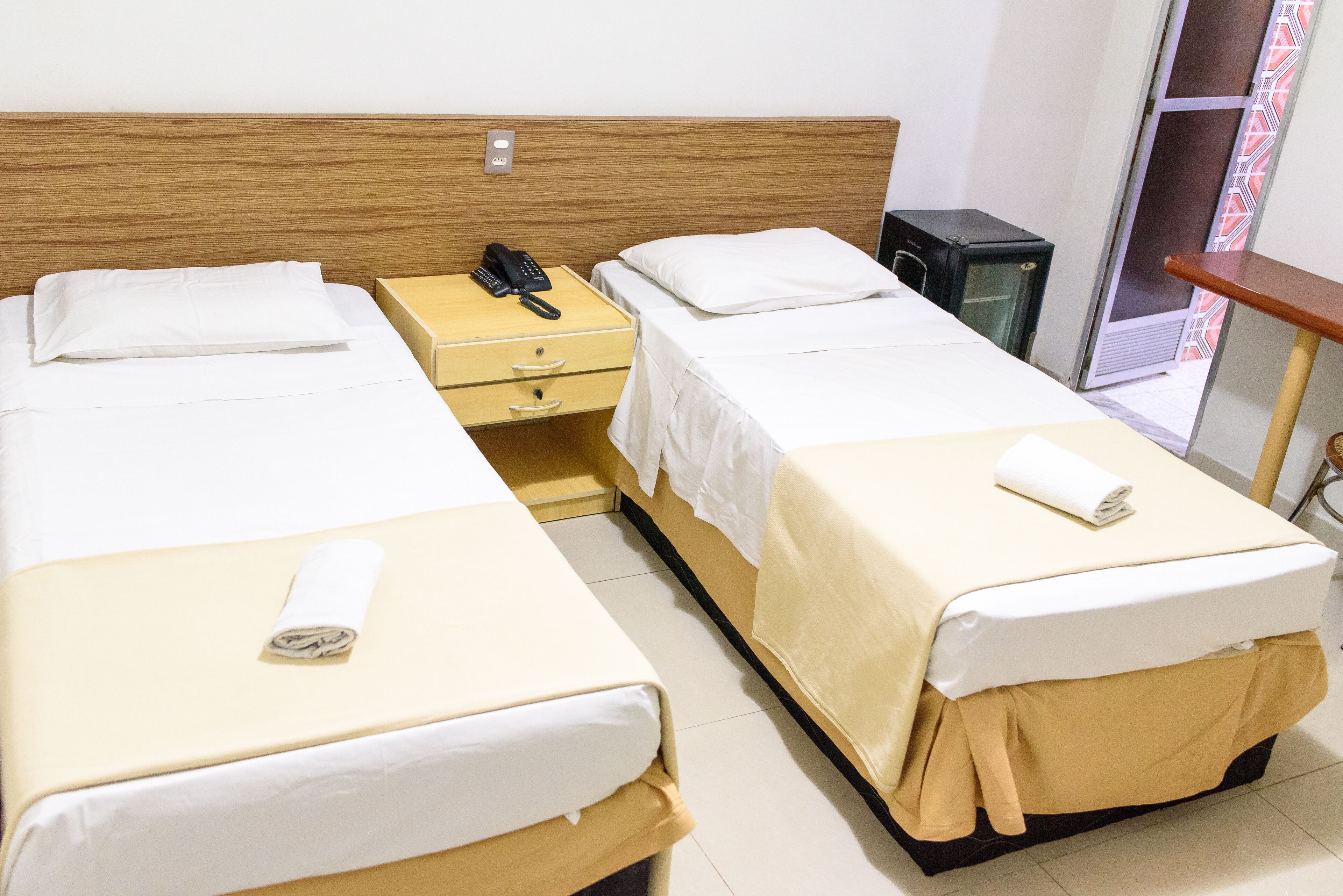 Quarto de hotel com duas camas de solteiro, mesa de cabeceira com telefone, pequeno armário e janela com vista.