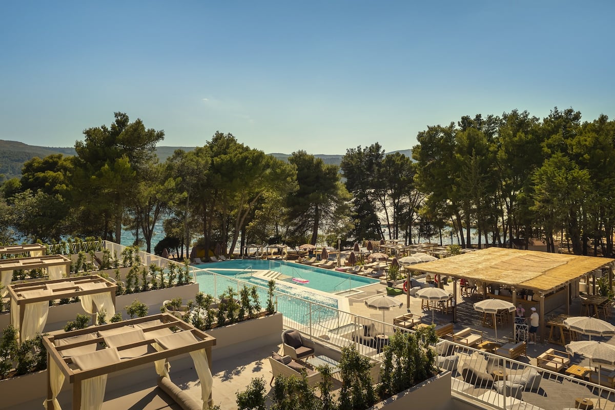 Área de piscina no terraço com cadeiras de descanso e guarda-sóis, cercada por árvores e com vista para um corpo d'água.