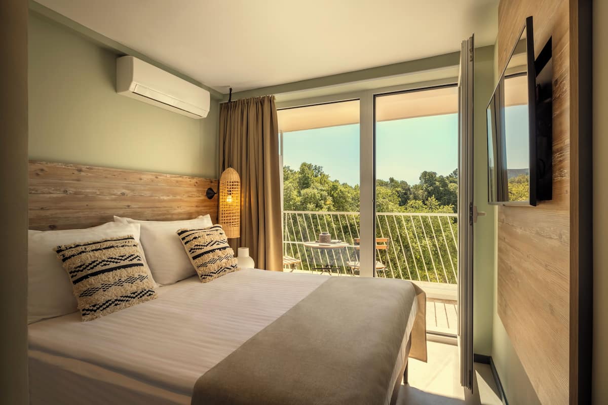 Quarto de hotel com cama grande, televisão de tela plana, varanda com mesa e cadeiras, e vista para o exterior.