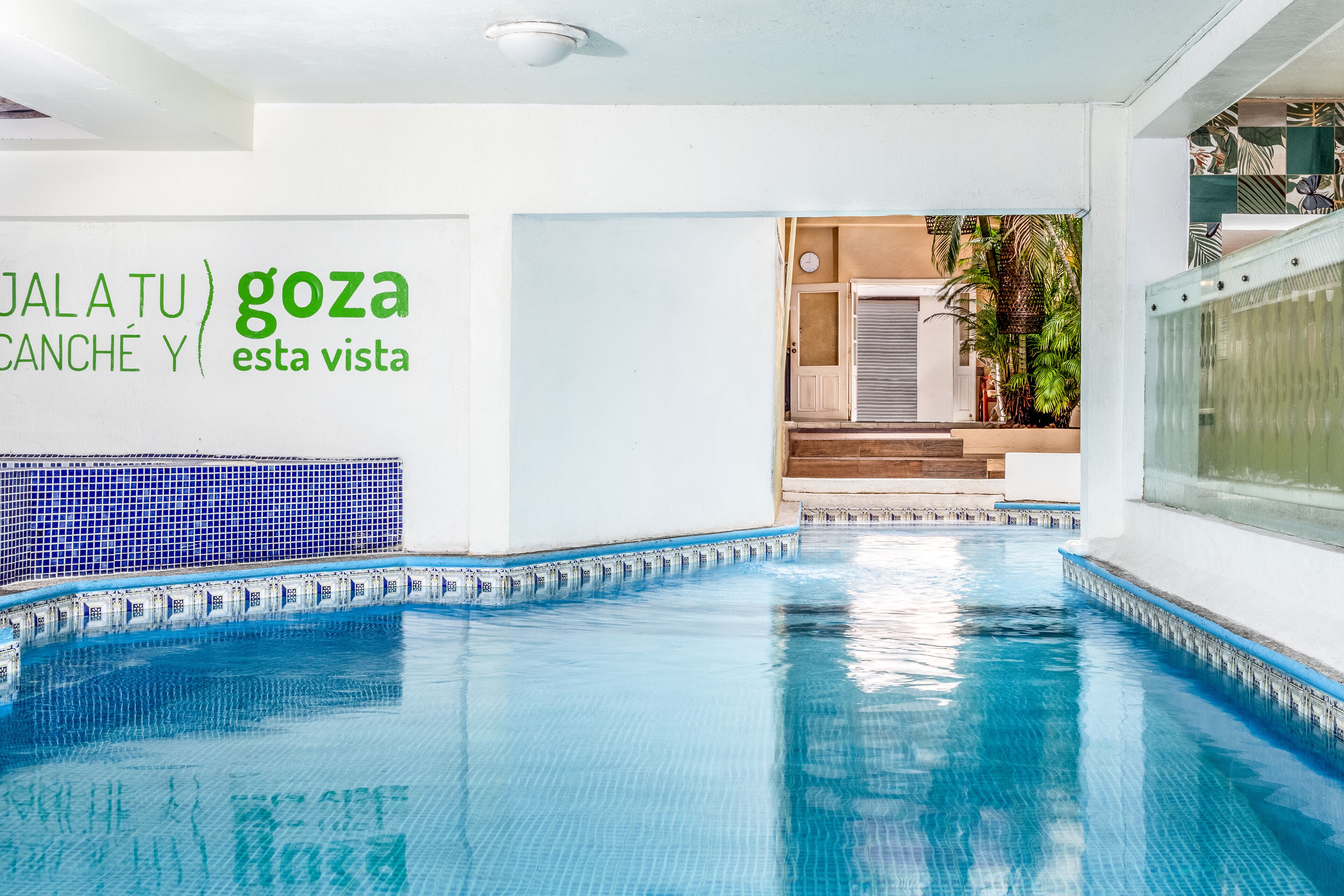 Piscina com borda azulejada, parede branca com uma placa, e vista de um edifício com porta e degraus.