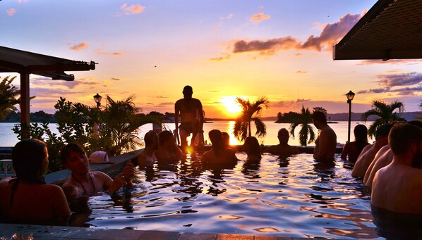 Um grupo de pessoas em uma piscina ao pôr do sol, com vista para a água e palmeiras.