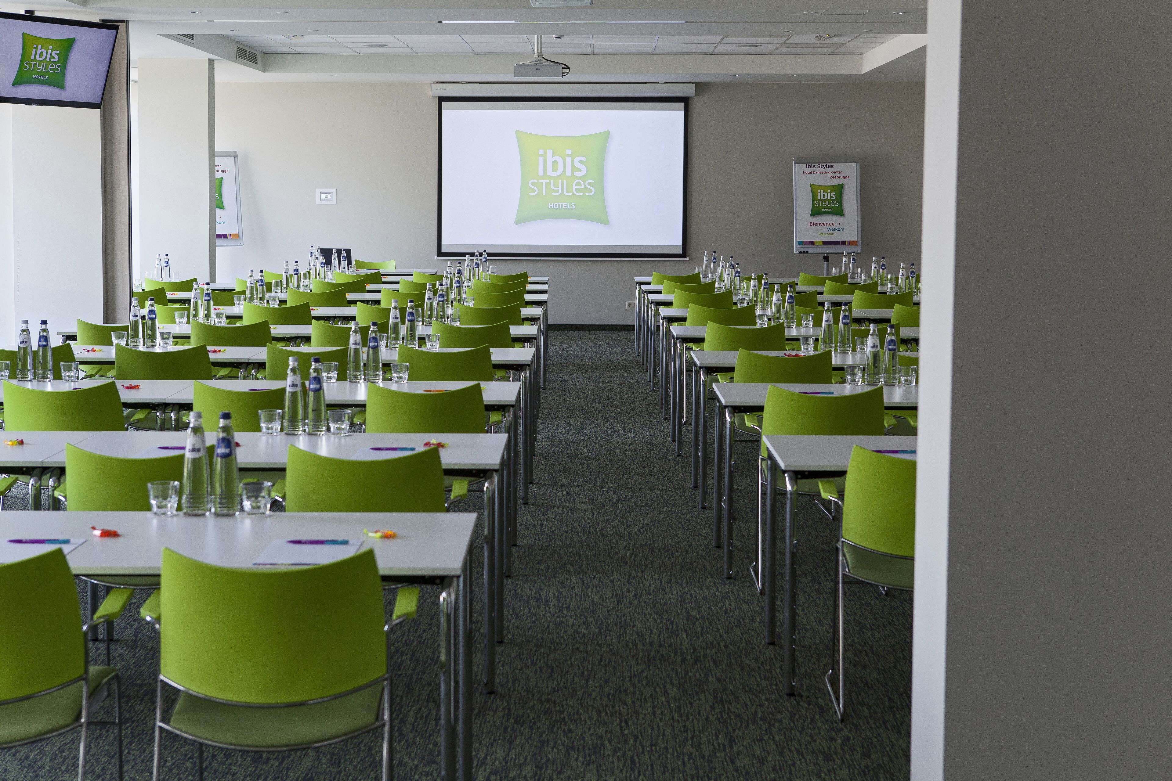 Uma sala de conferências com fileiras de mesas e cadeiras, uma tela de projetor exibindo 'ibis styles hotels' e uma televisão na parede.