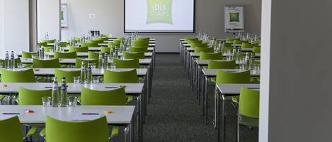 Uma sala de conferências com fileiras de mesas e cadeiras, uma tela de projetor exibindo 'ibis styles hotels' e uma televisão na parede.