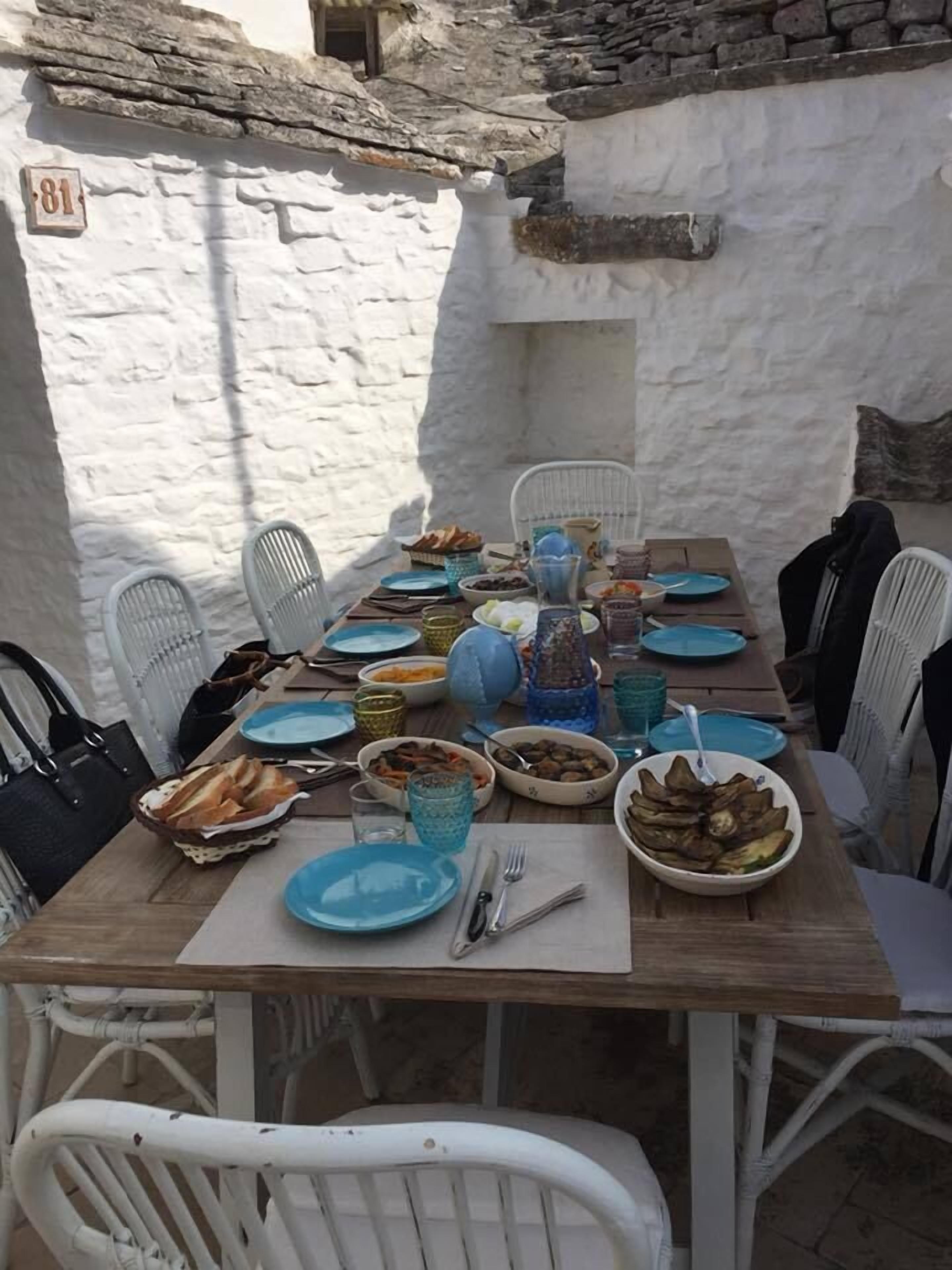 Mesa de jantar posta com diversos pratos e bebidas, cercada por cadeiras brancas, em ambiente rústico ao ar livre com paredes de pedra.