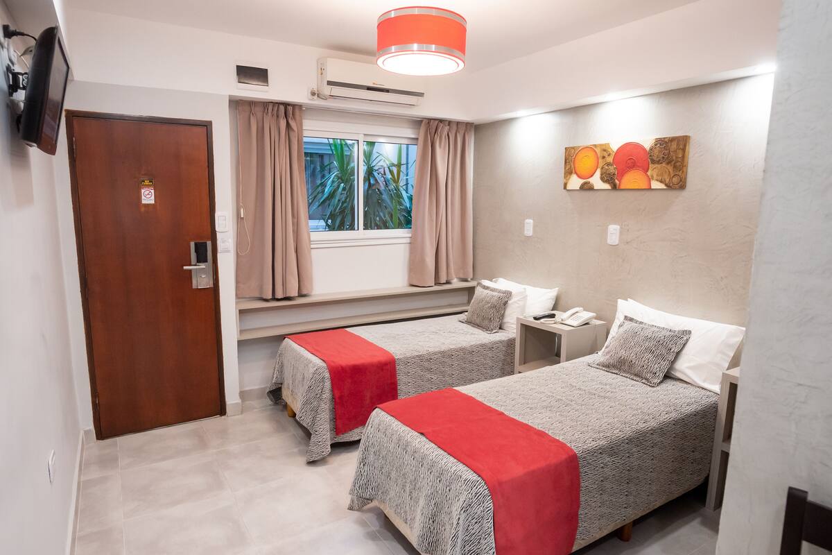 Quarto de hotel com duas camas, uma janela com cortinas, um quadro na parede e uma televisão montada na parede.