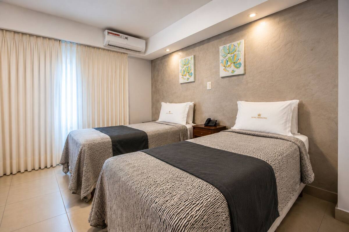Quarto de hotel com duas camas, uma janela com cortinas, um ar condicionado e dois quadros pendurados na parede.