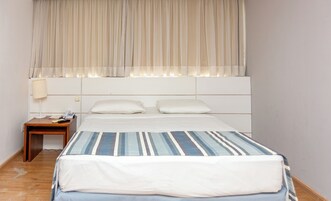 Quarto com cama, duas almofadas, mesa de cabeceira com abajur e criado-mudo com telefone.