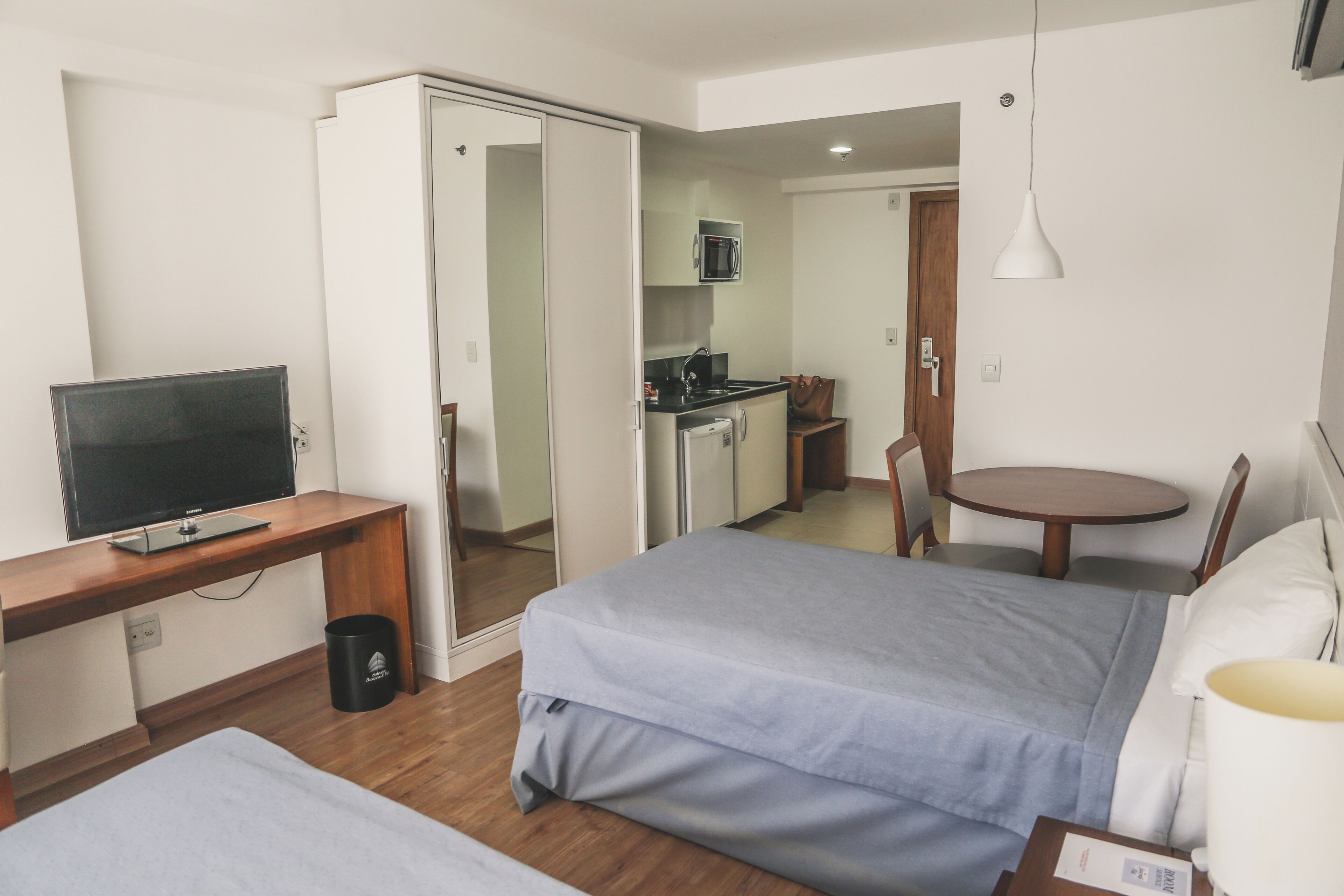 Quarto de hotel com cama, escrivaninha com televisão, pequena cozinha e área de refeições com mesa e cadeiras.