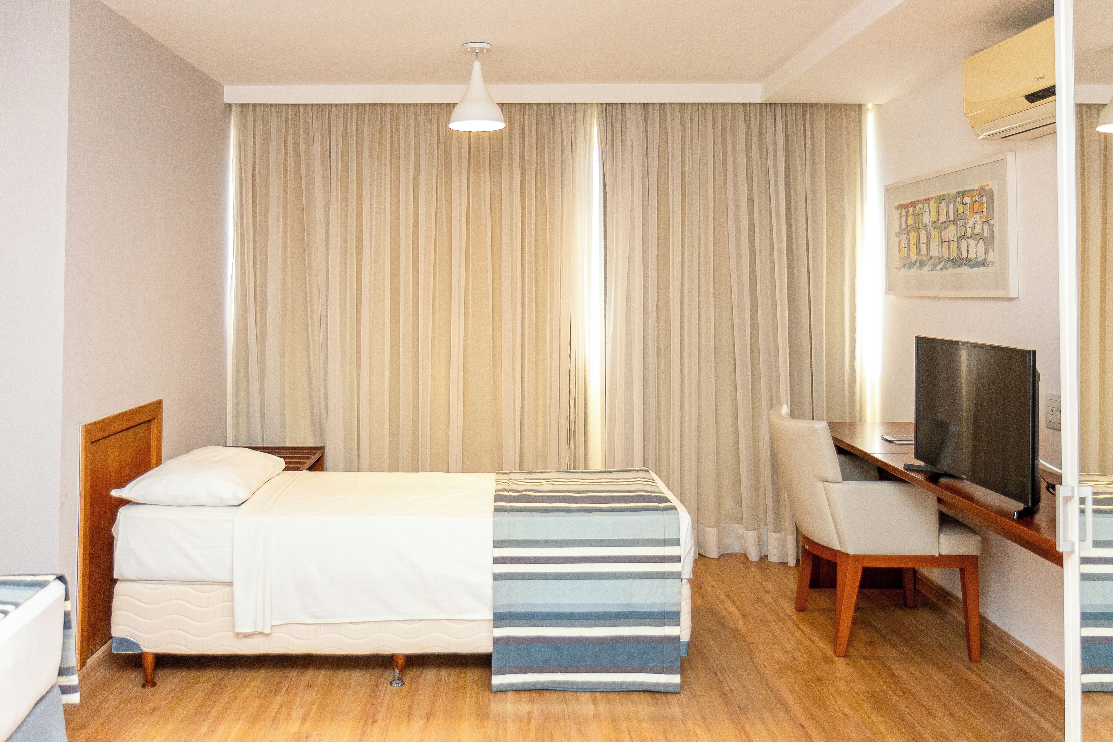 Quarto de hotel com cama, escrivaninha com televisão, uma cadeira e janela com cortinas.
