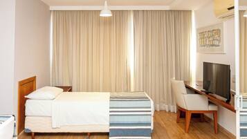 Quarto de hotel com cama, escrivaninha com televisão, uma cadeira e janela com cortinas.