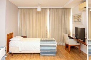 Quarto de hotel com cama, escrivaninha com televisão, uma cadeira e janela com cortinas.