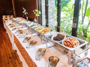 Disposição de buffet com diversos pratos e utensílios em bandejas, dispostos sobre uma mesa com vista para o verde lá fora.