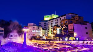Vista urbana noturna com edifícios iluminados, uma fonte e uma estrutura de madeira tradicional.