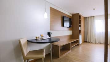 Quarto de hotel moderno com piso de madeira, uma mesa redonda com uma cadeira, uma TV fixada na parede e uma grande janela com cortinas.