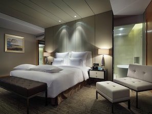 Quarto de hotel moderno com uma cama grande, uma mesa de cabeceira com abajur, um banco e uma cadeira.