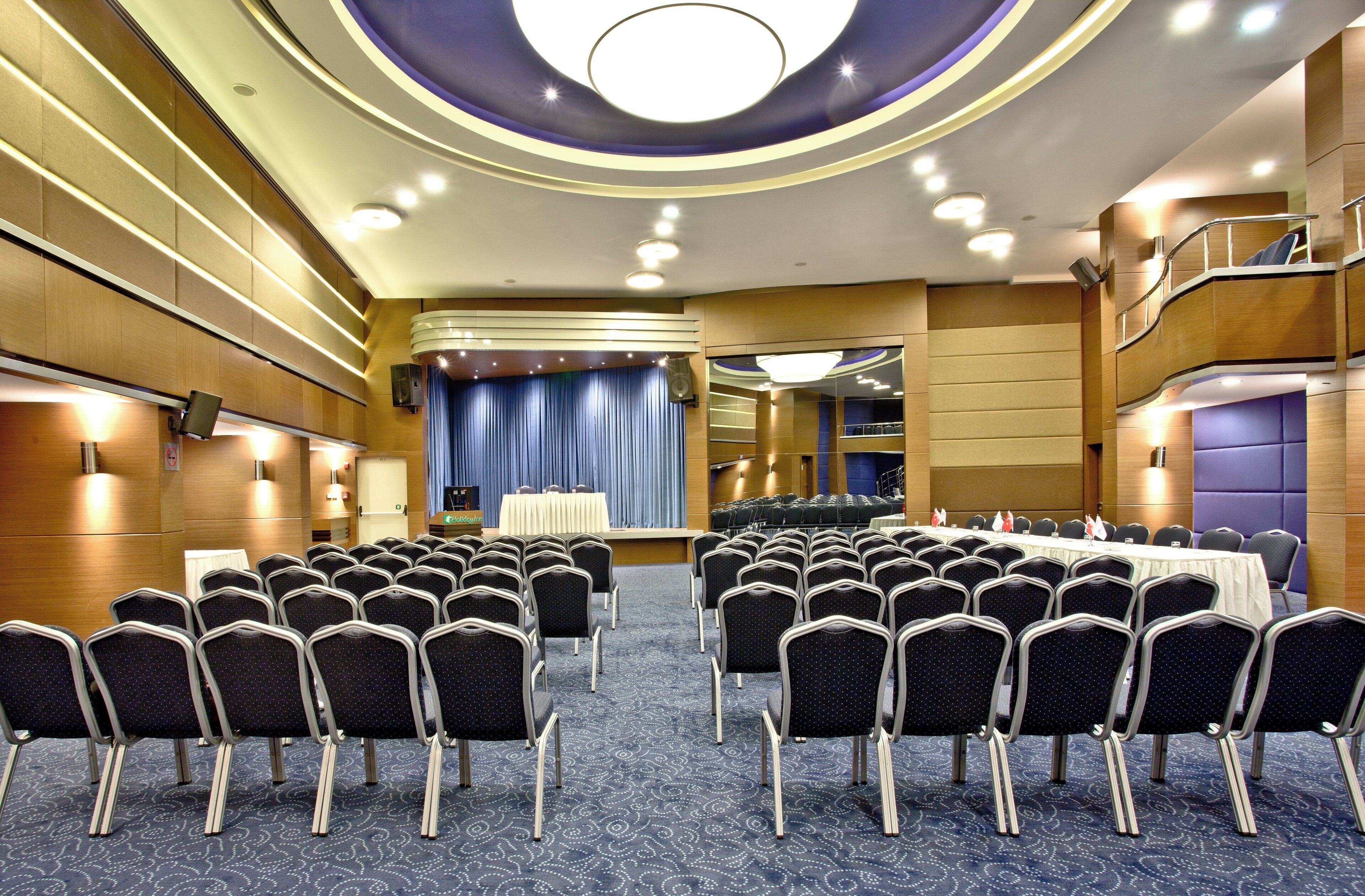 Uma sala de conferências com fileiras de cadeiras, um palco com cortinas e uma varanda.