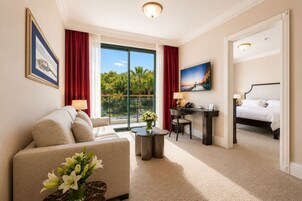 Quarto de hotel com sofá, escrivaninha, cama, televisão e uma grande janela com cortinas.