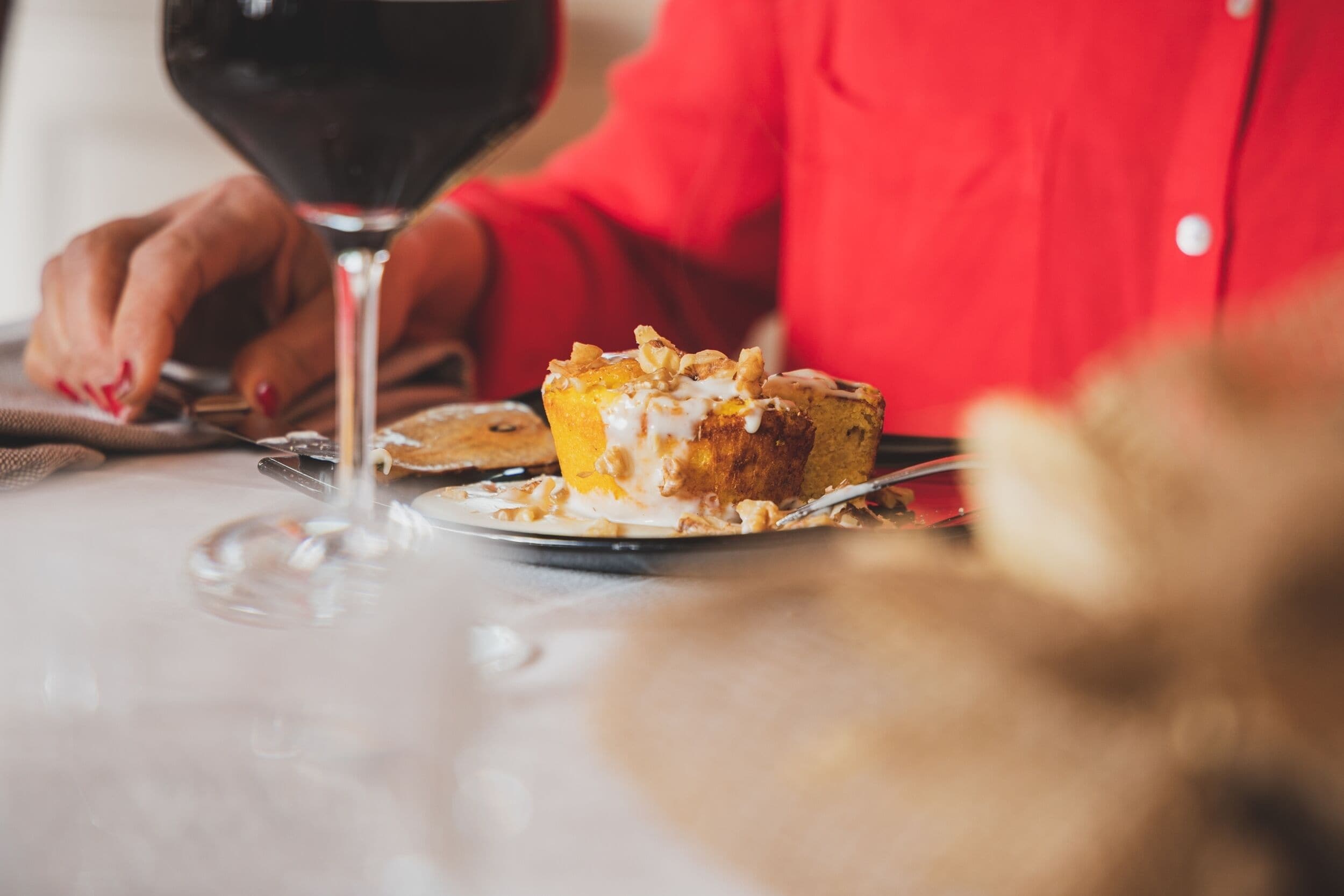 Uma pessoa vestindo uma camisa vermelha sentada à mesa com um copo de vinho tinto e um prato com sobremesa.