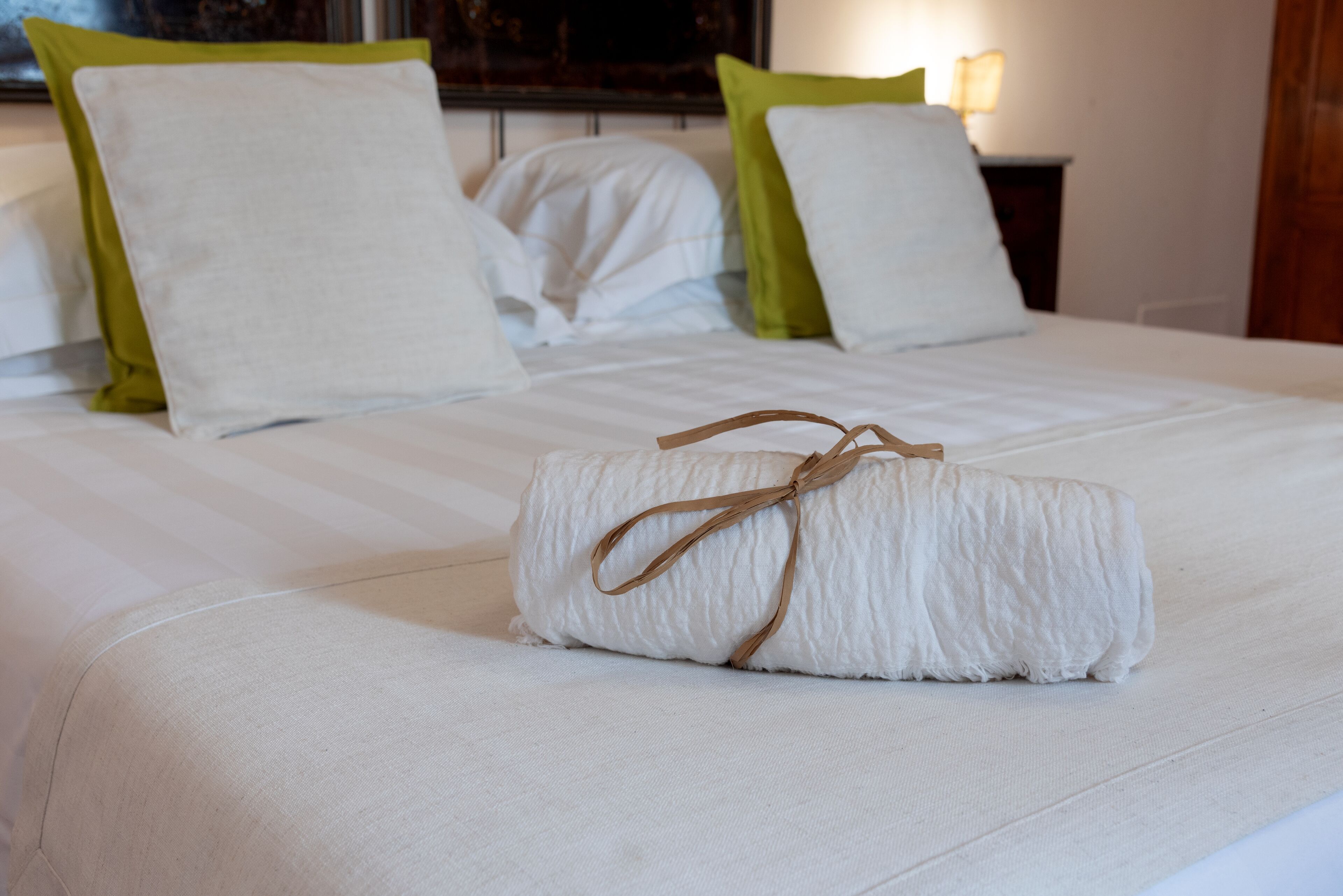Cama bem arrumada com lençóis brancos e uma toalha enrolada amarrada com um laço de fita marrom.
